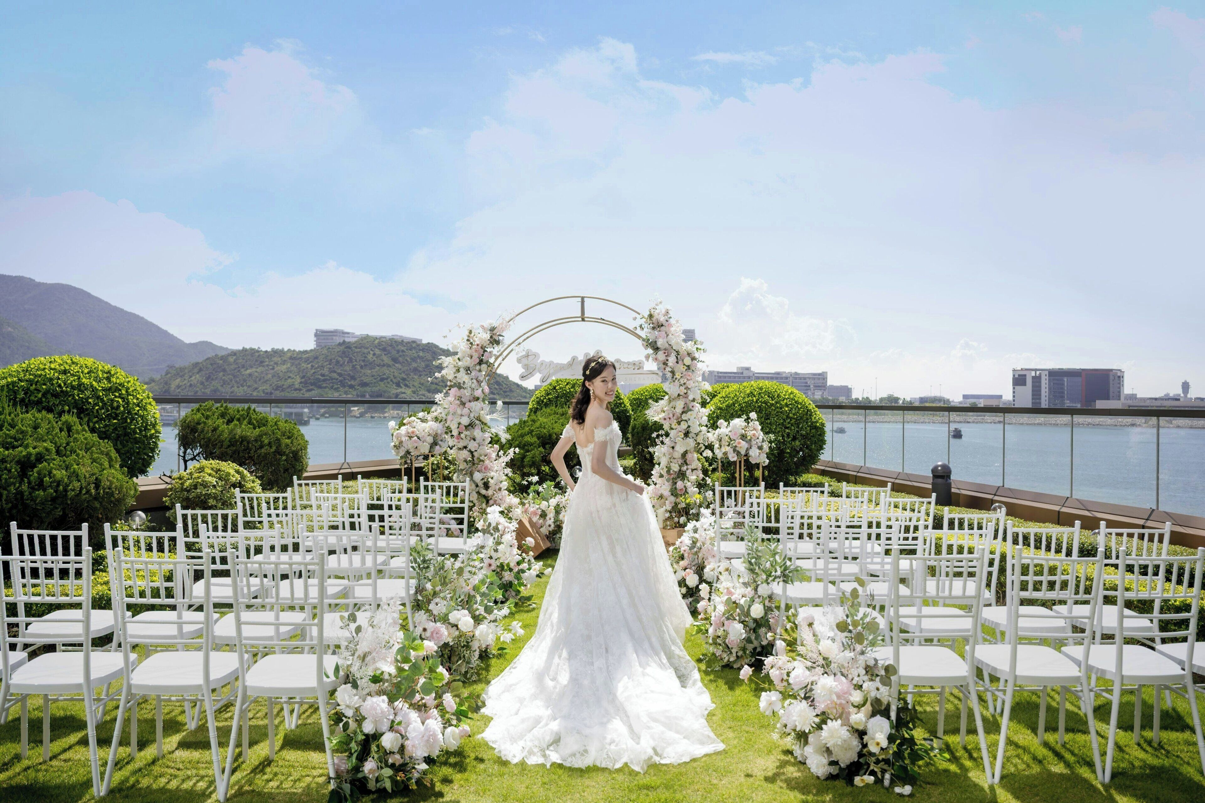 Preparação para cerimônia de casamento com cadeiras brancas e decoração floral, com vista para um corpo d'água e colinas.