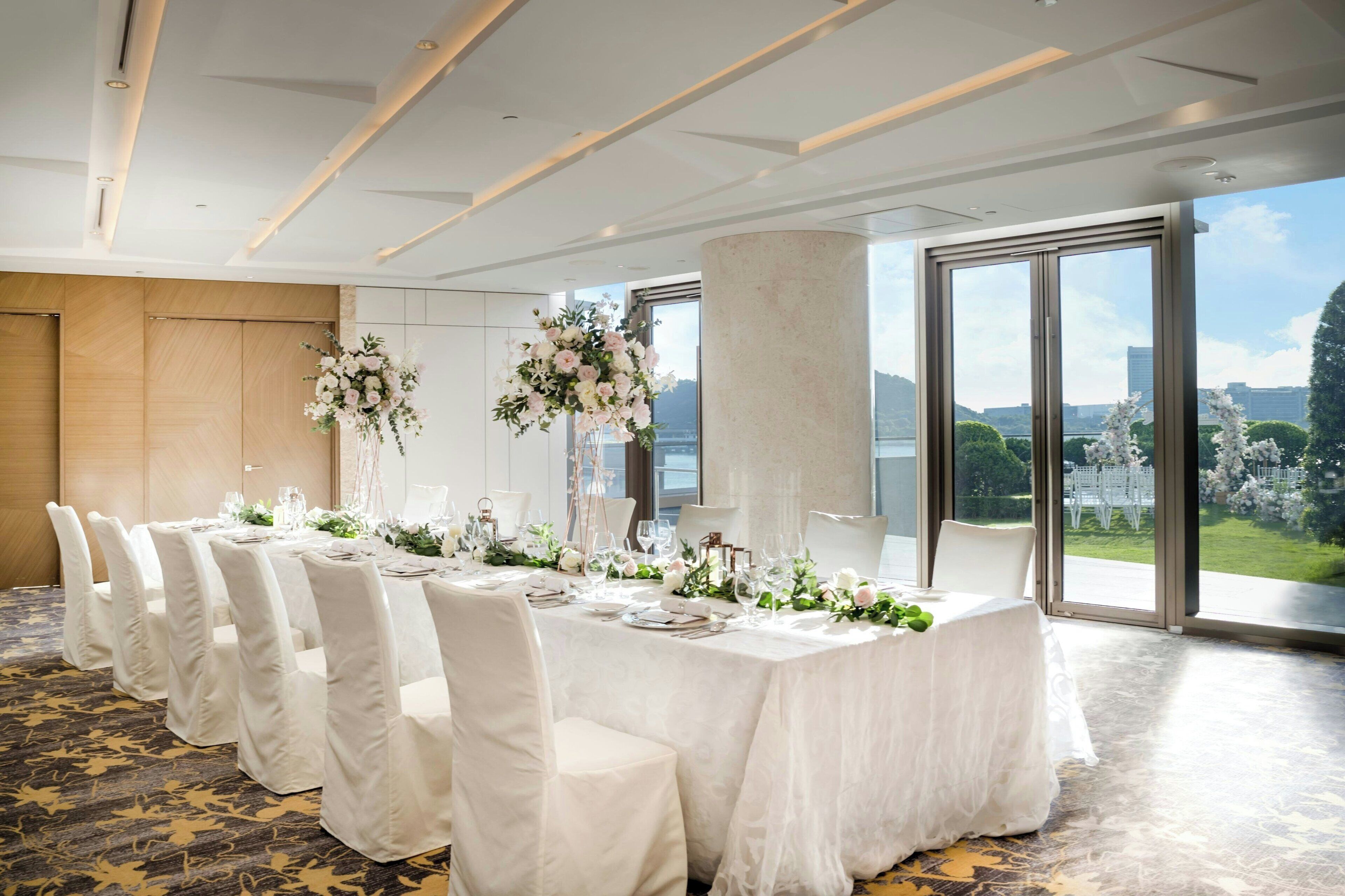 Uma sala de banquetes com uma longa mesa preparada para um evento formal, amplas janelas com vista para um lago e horizonte urbano, e um design de interiores moderno.