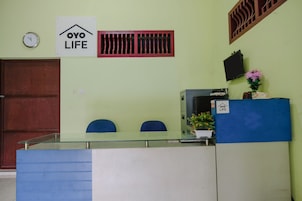 Uma área de recepção com balcão de vidro, duas cadeiras azuis, um relógio, uma ventilação, uma TV e um aviso com a inscrição "OYO LIFE".