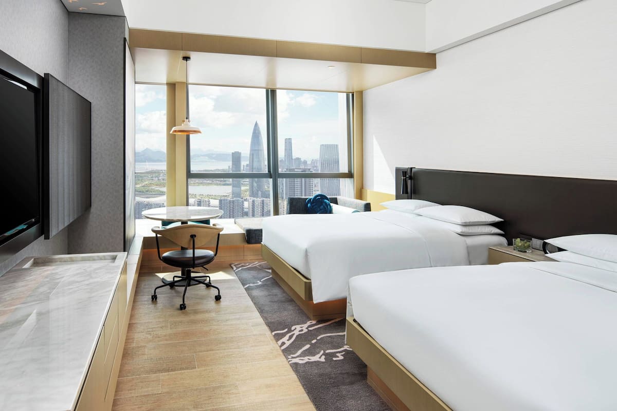 Quarto de hotel com duas camas, uma escrivaninha, uma cadeira, televisão e uma janela grande com vista para a cidade.