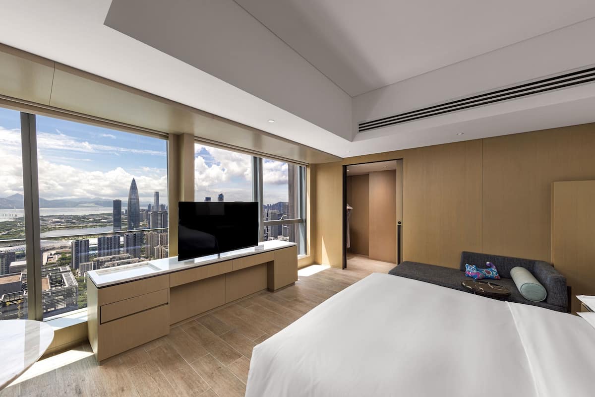 Quarto de hotel moderno com uma cama grande, uma televisão em uma escrivaninha e vista para a cidade através de amplas janelas.