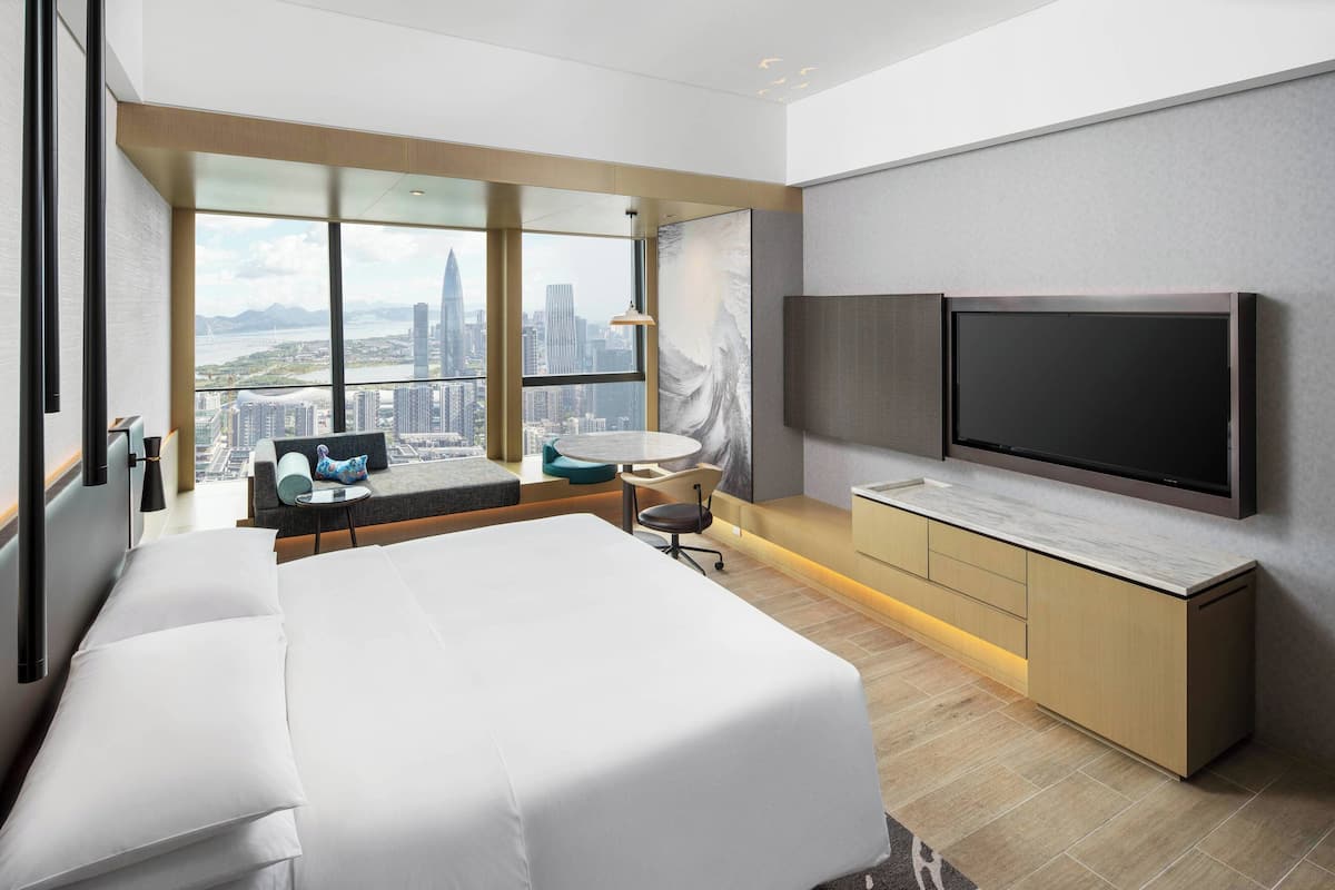 Quarto de hotel moderno com uma cama grande, escrivaninha, cadeira, televisão e vista para a cidade.
