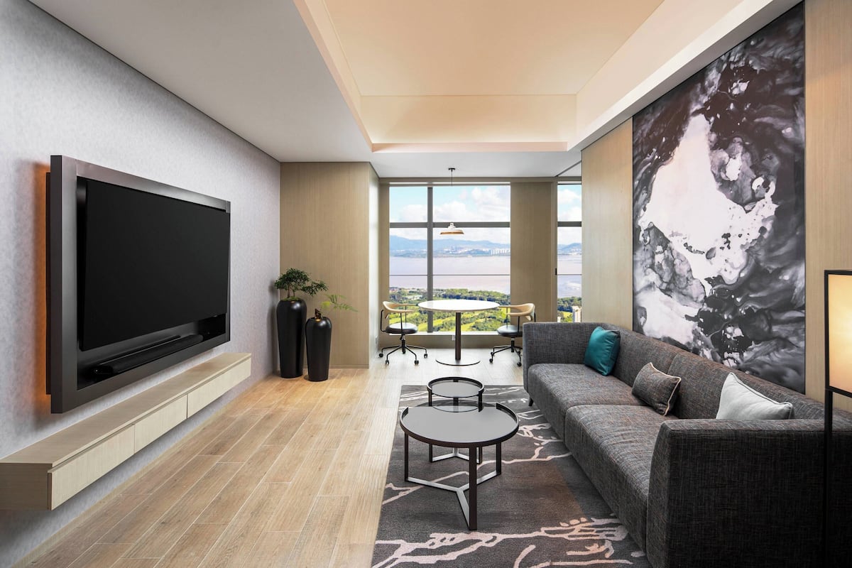 Uma sala de estar moderna com uma grande TV de tela plana, um sofá cinza com almofadas verde-água, uma mesa de centro e vista para um horizonte urbano através de amplas janelas.