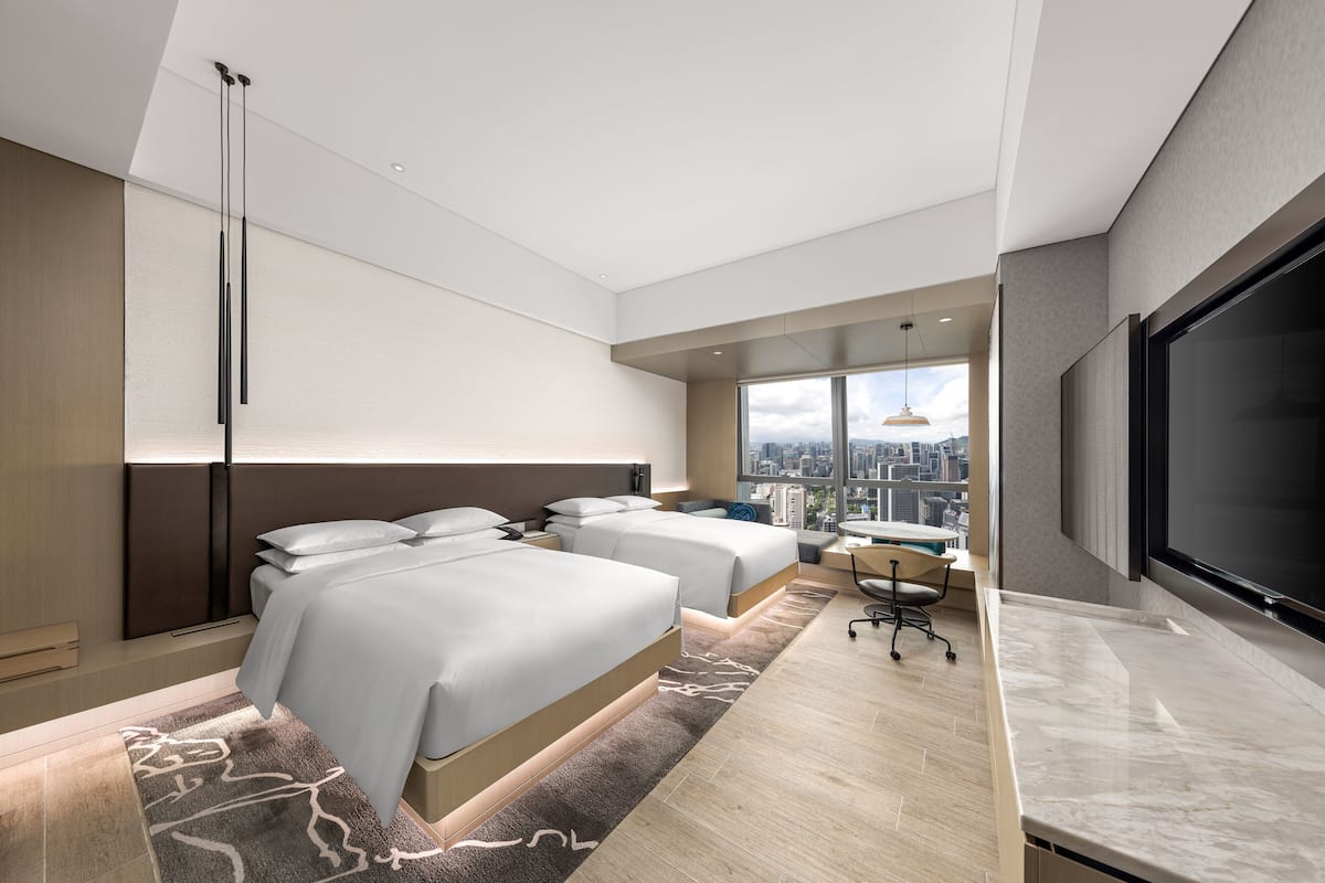 Quarto de hotel moderno com duas camas, uma escrivaninha, uma janela grande com vista para a cidade e uma televisão de tela plana.