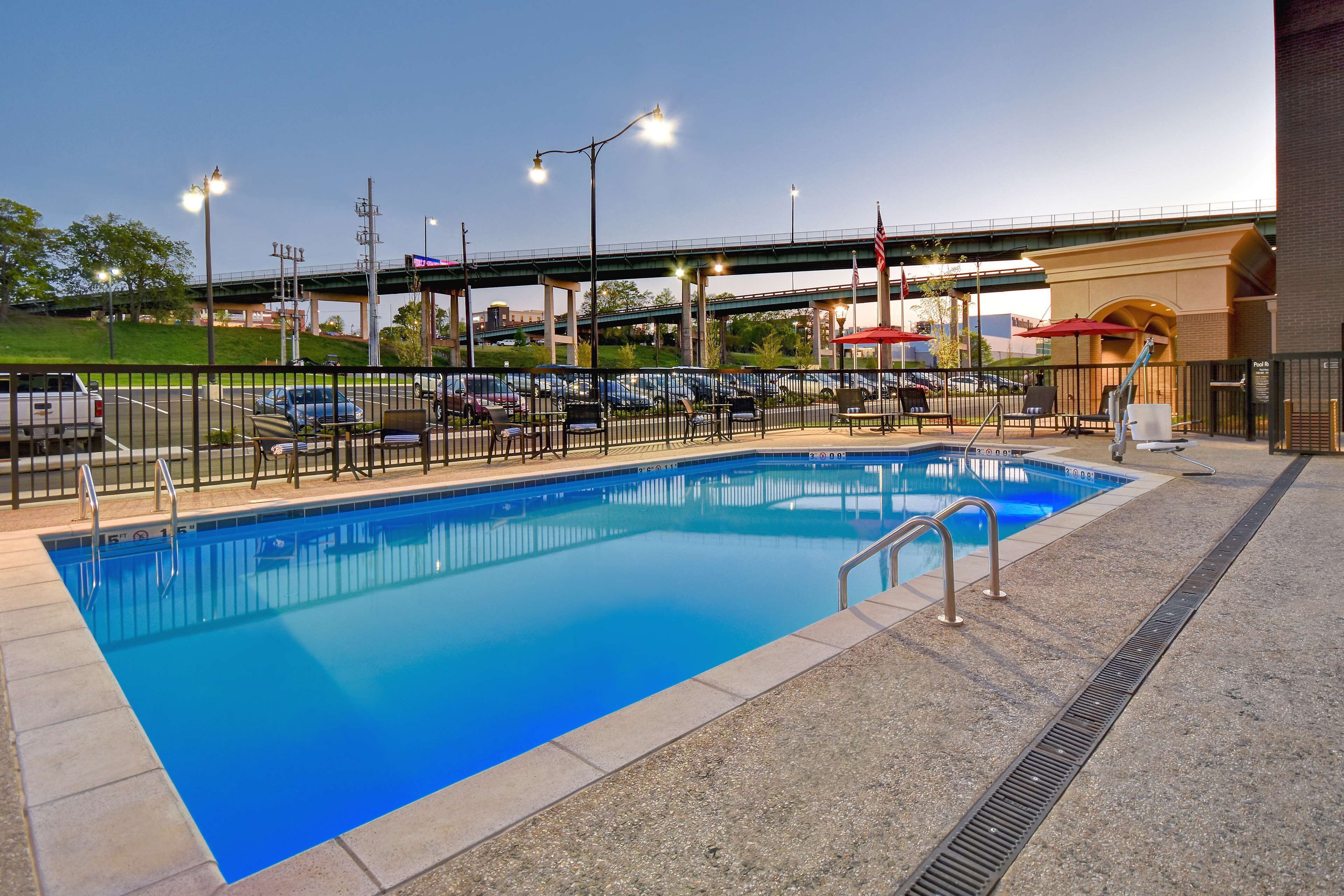 Piscina com deck pavimentado, guarda-corpos de metal e vista para um estacionamento e uma ponte ao fundo.