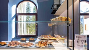 Buffet de café da manhã com diversos pães e pastéis expostos em uma vitrine de vidro, com aviso sobre medidas de distanciamento social.
