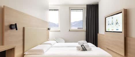 Quarto de hotel com duas camas, televisão na parede e vista para montanhas pelas janelas.