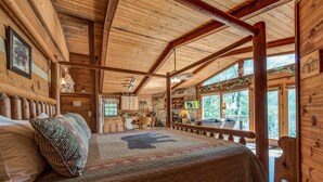 Interior de cabana de madeira com cama, paredes rústicas, área de jantar e grandes janelas.