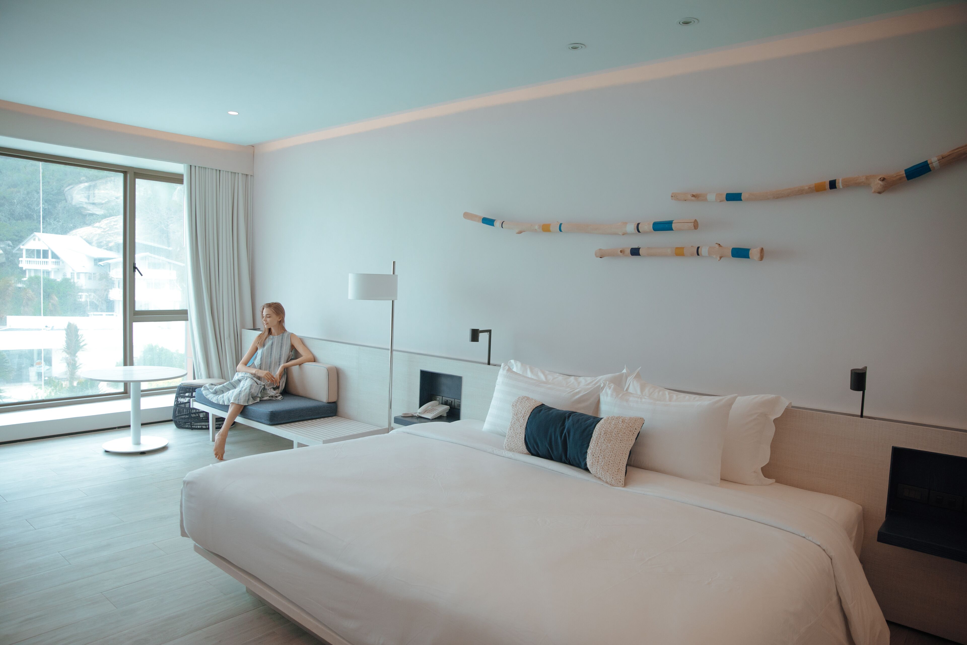 Quarto de hotel moderno com uma cama grande, um espaço de estar com sofá e mesa, mesa de cabeceira com abajur e vista através de uma janela ampla.