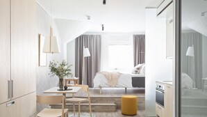 Quarto de hotel moderno com cama, mesa pequena com cadeiras, sofá, área de cozinha e mesa de jantar com um vaso de flores.