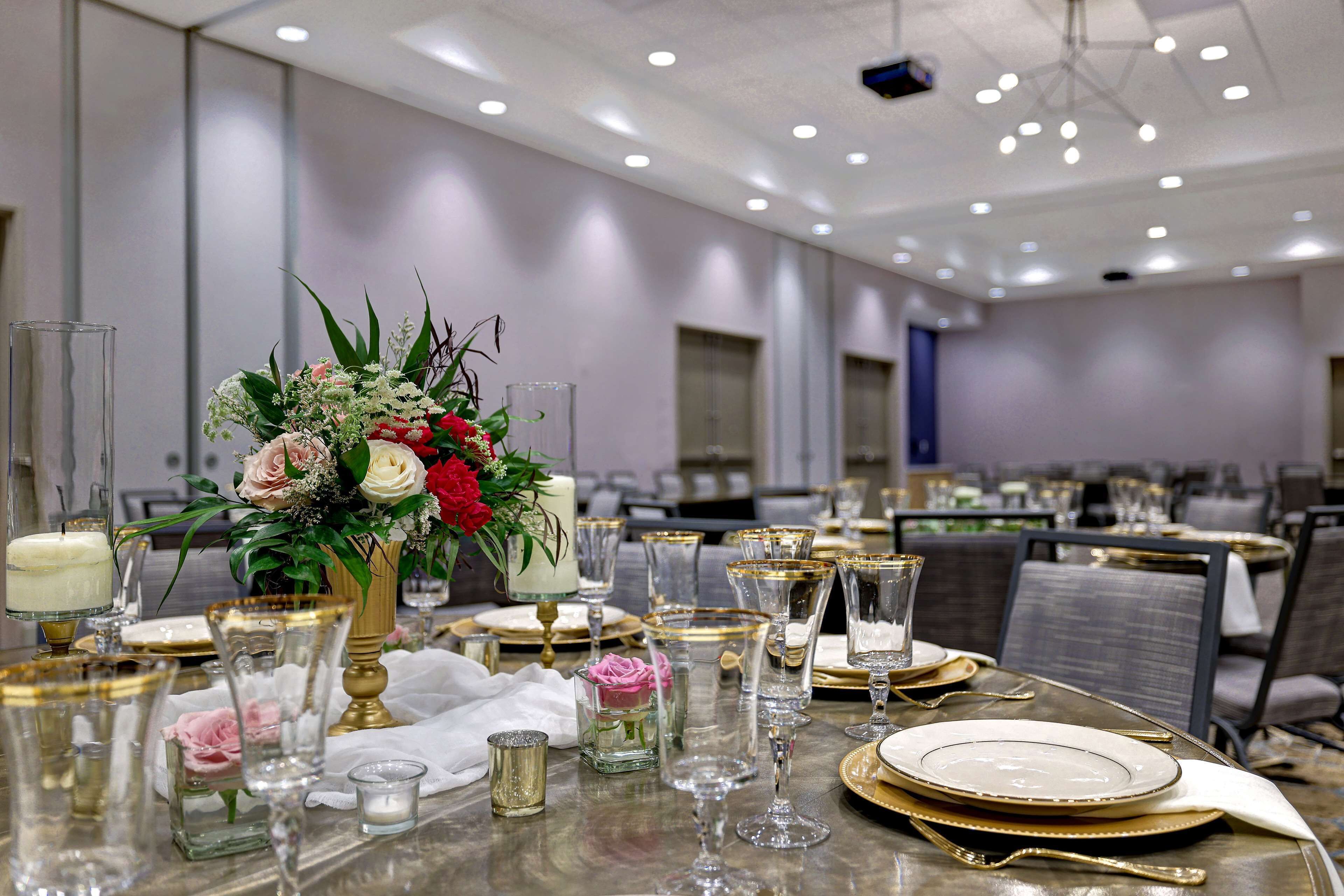 Uma sala de banquetes com mesas montadas para um evento formal, com copos, pratos e um arranjo central com flores e velas.