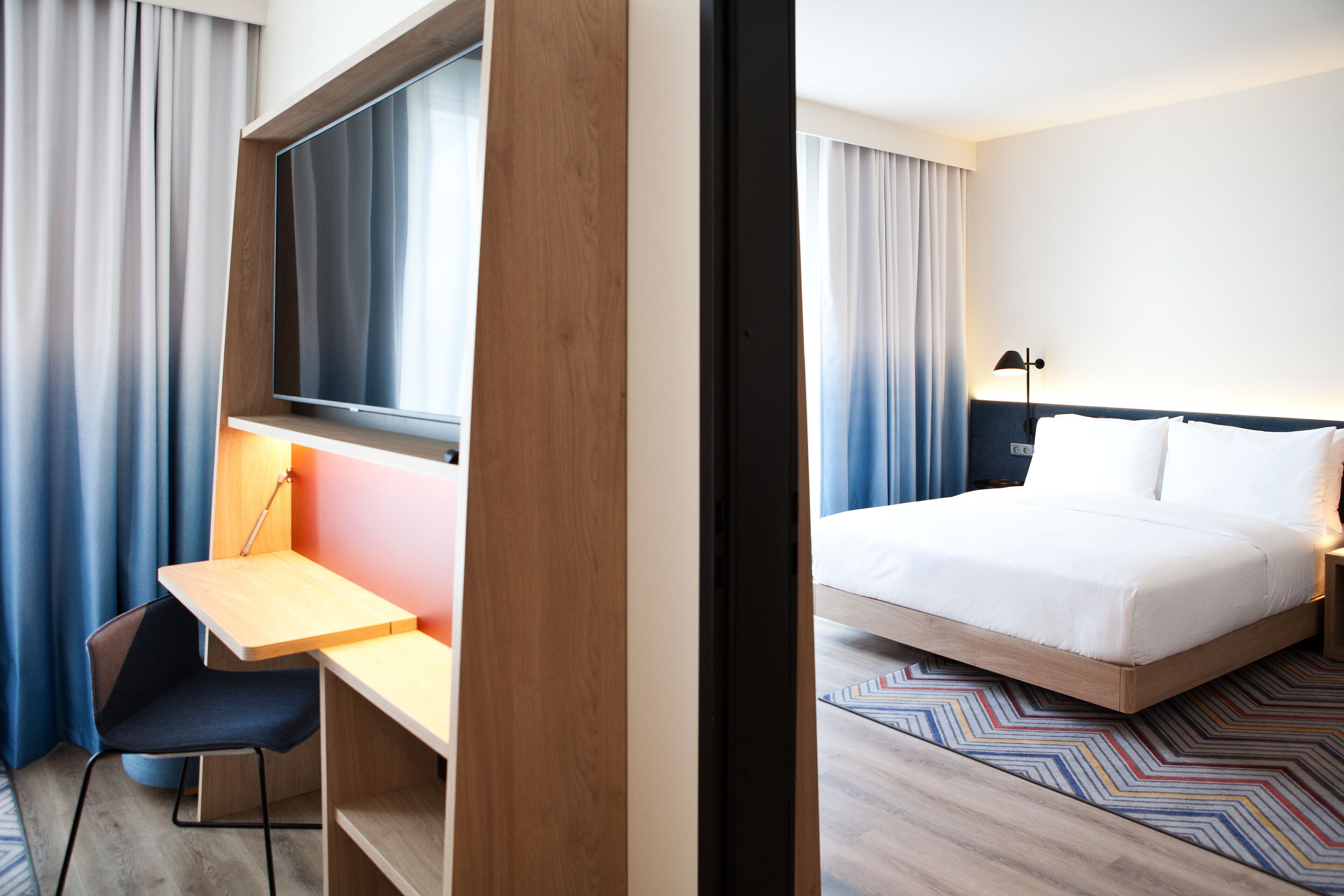 Quarto de hotel com cama, escrivaninha, cadeira, televisão e luminária.