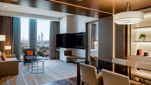 Quarto de hotel moderno com televisão grande, assentos confortáveis, área de refeições e vista para a cidade.