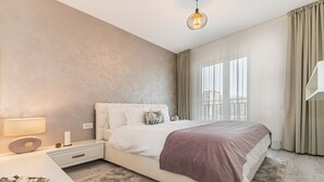 Quarto moderno com uma cama grande, mesas de cabeceira, uma luminária e vista para a cidade pela janela.
