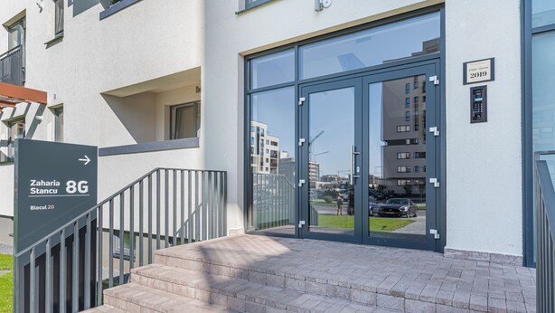 Entrada de um edifício moderno com portas de vidro, uma placa indicando 'Zaharia Stancu 86' e 'Blocul 20', e uma escadaria que leva à entrada.