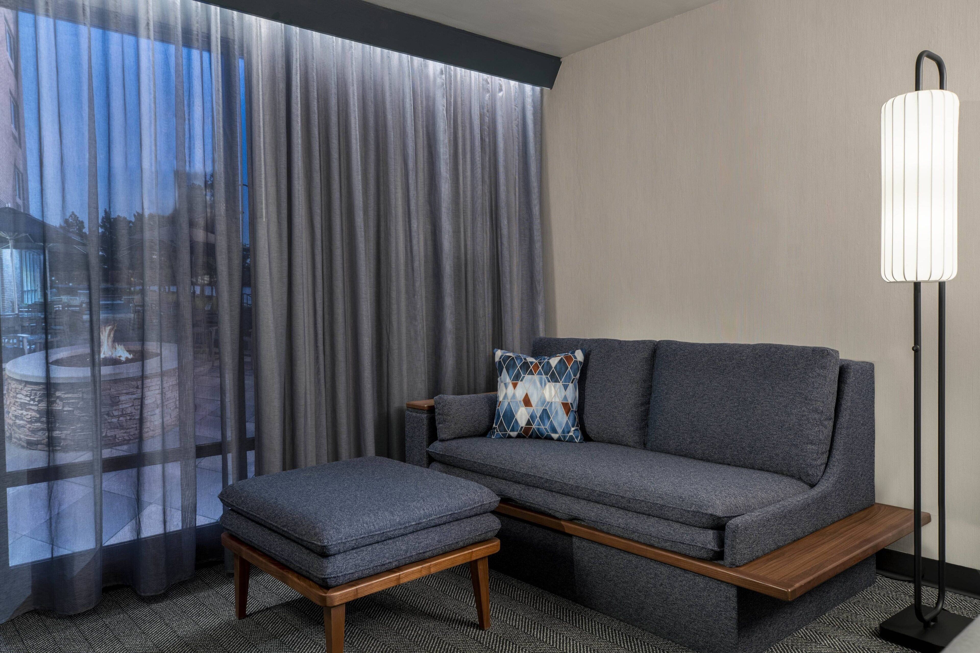 Quarto de hotel moderno com sofá cinzento, um banquinho coordenado, abajur e uma grande janela com cortinas translúcidas.