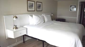 Cama bem arrumada com lençóis brancos e cabeceira, mesa de cabeceira com abajur, e duas fotos emolduradas na parede.