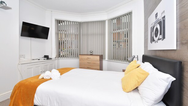 Quarto bem arrumado com cama, mesa de cabeceira, TV fixada na parede e janela com persianas.