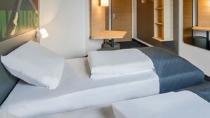Quarto de hotel com duas camas, uma mesa pequena, um espelho e um prateleira na parede.