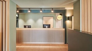 Balcão de recepção com design moderno, com acabamento em madeira clara e divisória de vidro. Dois recepcionistas estão atrás do balcão, e uma placa acima deles diz "B&B Hotels".