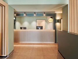 Balcão de recepção com design moderno, com acabamento em madeira clara e divisória de vidro. Dois recepcionistas estão atrás do balcão, e uma placa acima deles diz "B&B Hotels".