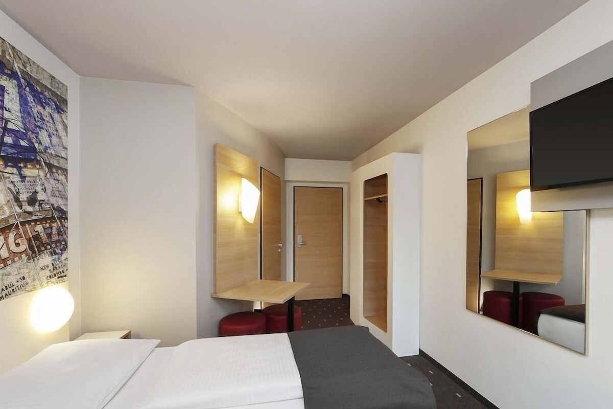 Quarto de hotel com uma cama, escrivaninha, cadeira, guarda-roupa e televisão.