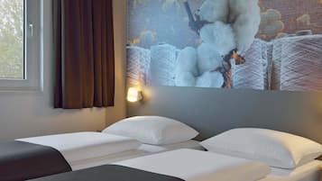 Quarto de hotel com duas camas de solteiro, janela com cortinas, luminária fixada na parede e uma grande obra de arte com tema de algodão.
