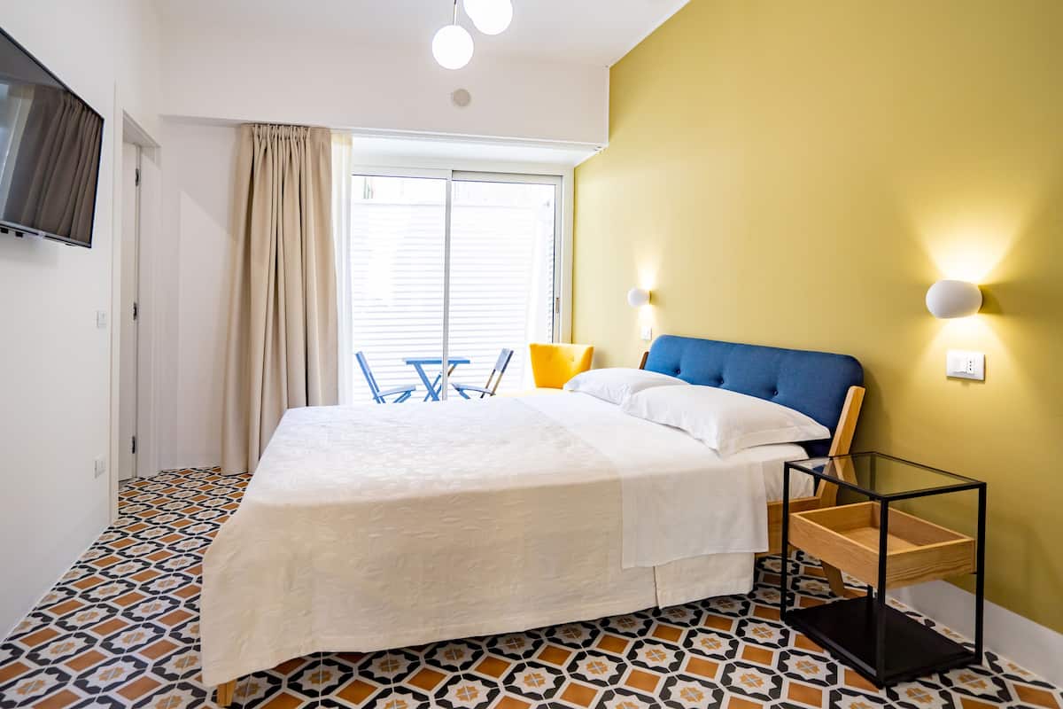 Quarto com cama, mesa de cabeceira, cadeira, janela com cortinas e televisão na parede.