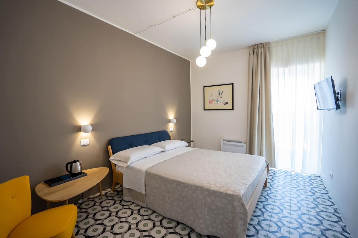 Quarto de hotel com cama, uma cadeira, mesa com chaleira, televisão e janela com cortinas.