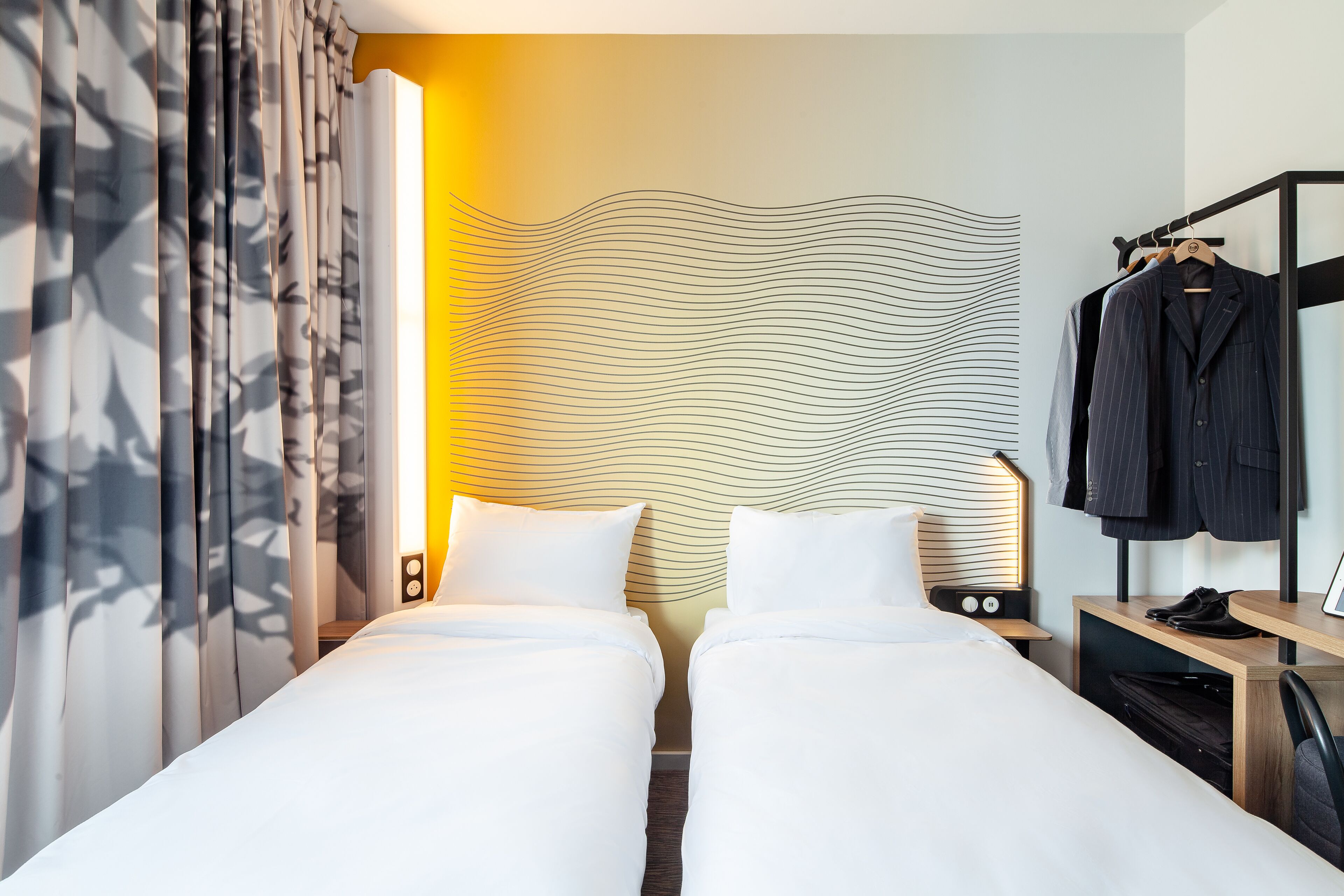 Quarto de hotel com duas camas, cabeceira com design ondulado, mesa de cabeceira com abajur e um cabideiro com um terno.