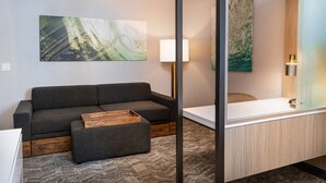 Quarto de hotel moderno com sofá cinzento, mesa de centro de madeira, pintura abstrata grande, abajur e divisória de vidro.