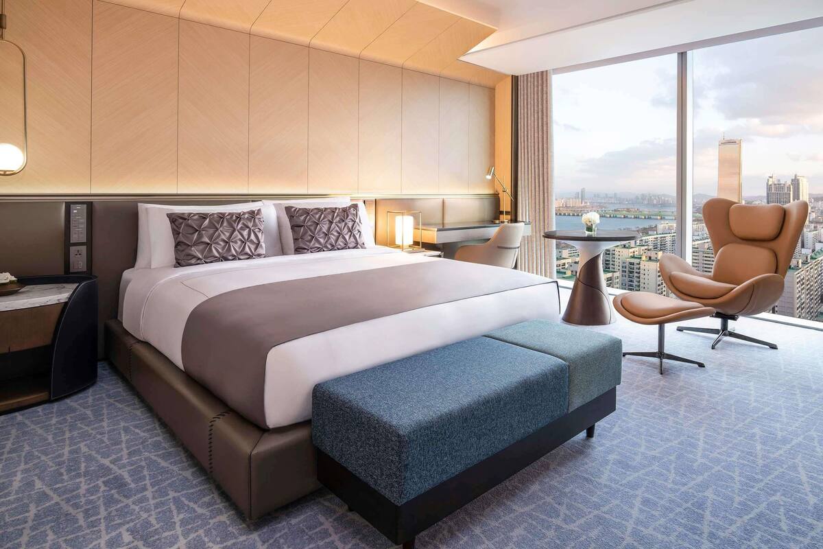 Quarto de hotel moderno com uma cama grande, área de estar com uma poltrona e banquinho, escrivaninha e uma janela ampla com vista para a cidade.