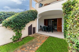 Uma área de estar coberta ao ar livre, com mesa e cadeiras, um caminho de pedra e vista para um edifício com varanda.