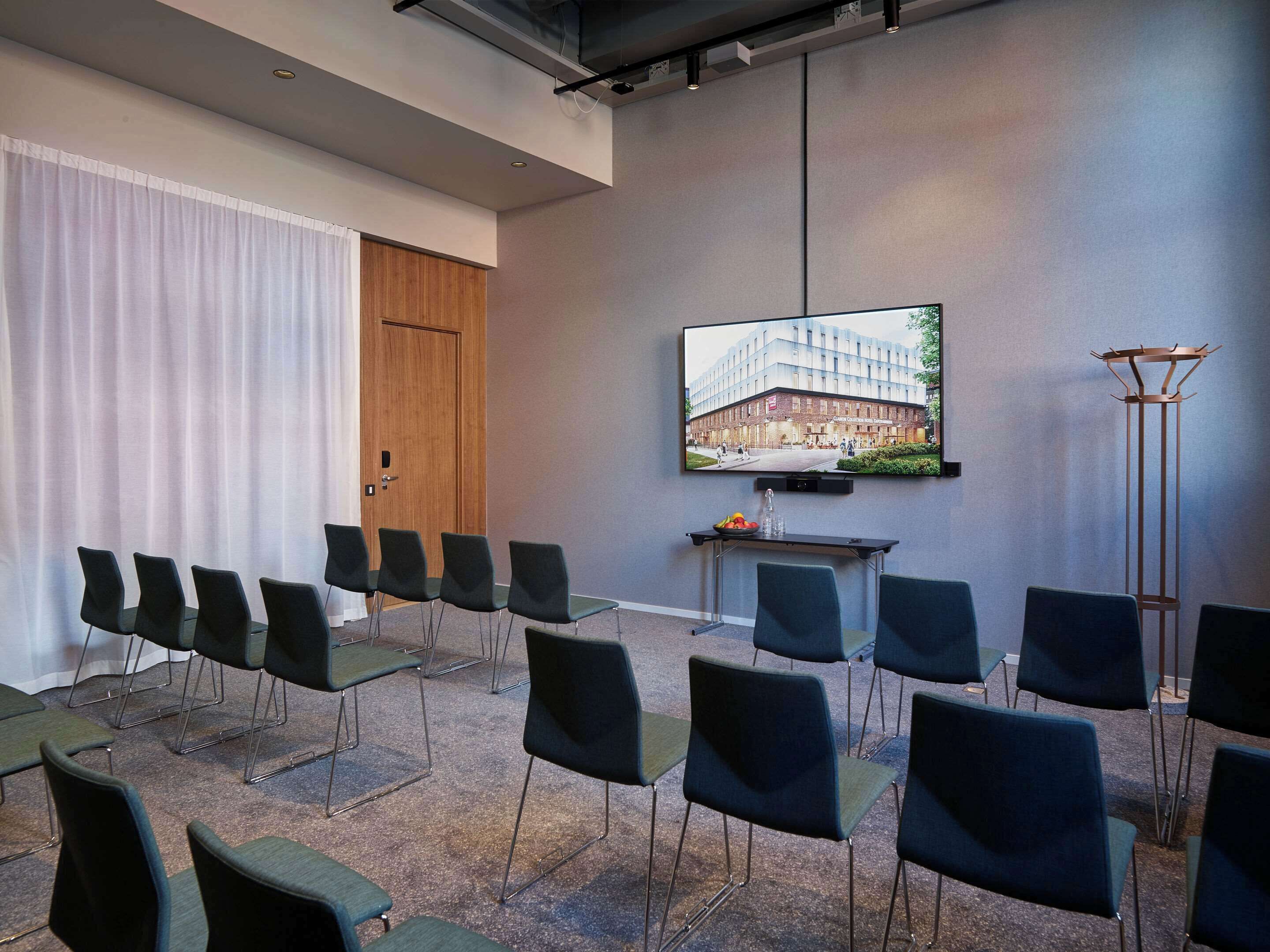 Uma sala de conferência com fileiras de cadeiras, uma tela de televisão, uma porta de madeira e um abajur.