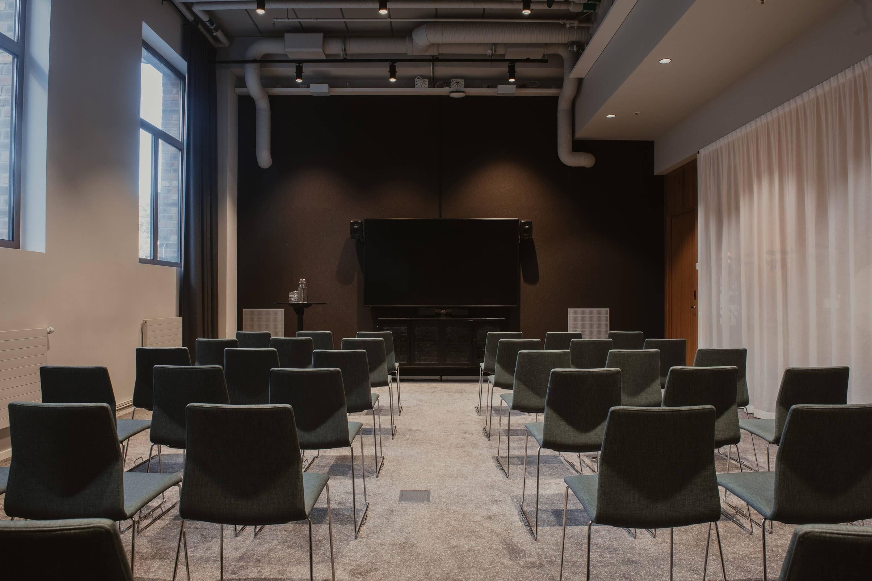 Uma sala de conferência com fileiras de cadeiras voltadas para um palco com uma tela grande, lareira e uma janela à esquerda.