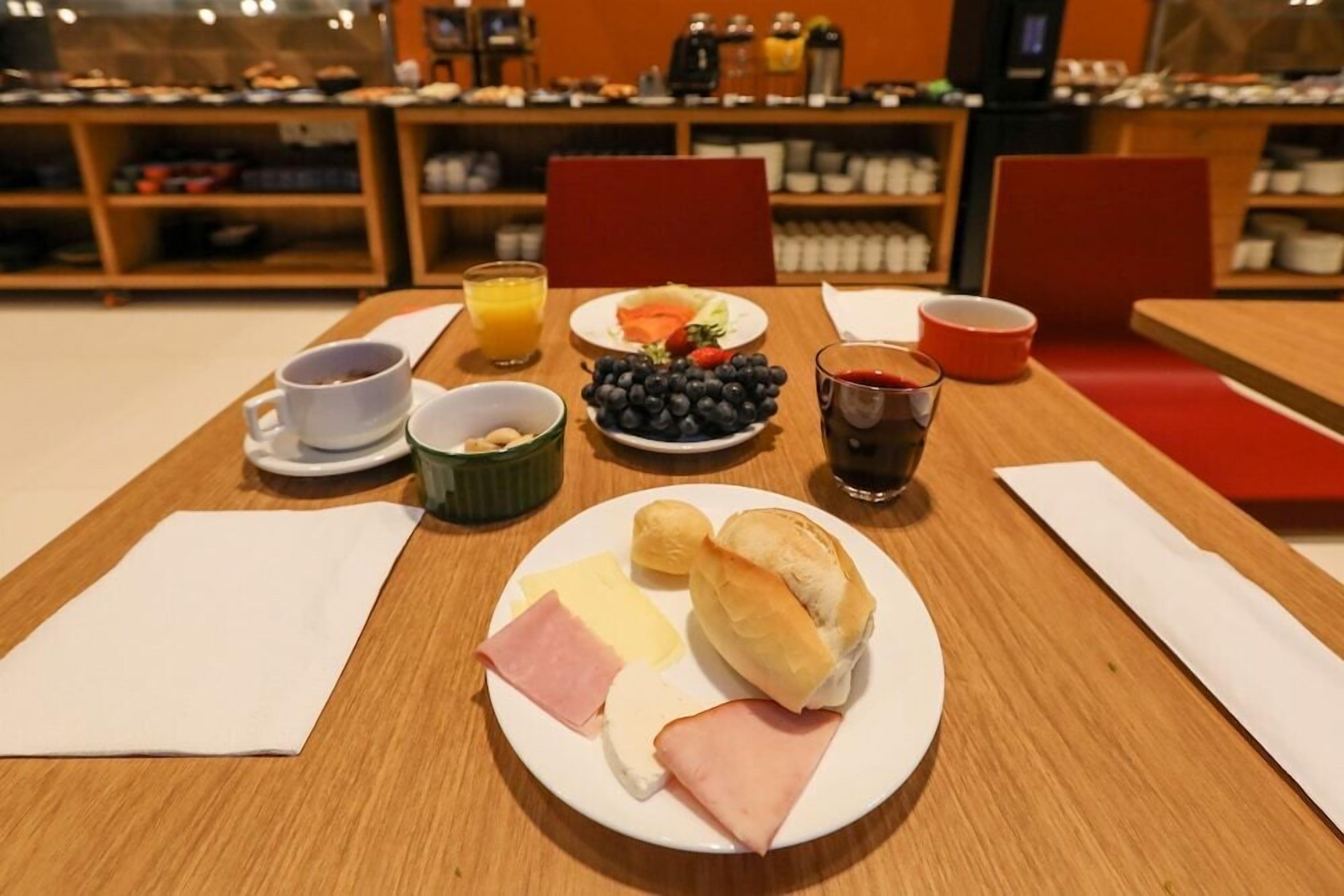 Mesa de café da manhã com pratos de comida, incluindo pão, queijo, presunto e frutas, além de xícaras de café e suco.