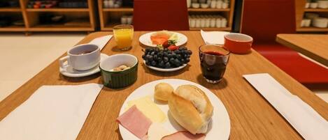 Mesa de café da manhã com pratos de comida, incluindo pão, queijo, presunto e frutas, além de xícaras de café e suco.