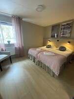 Quarto com duas camas, janela com cortinas, radiador e uma mesa pequena.