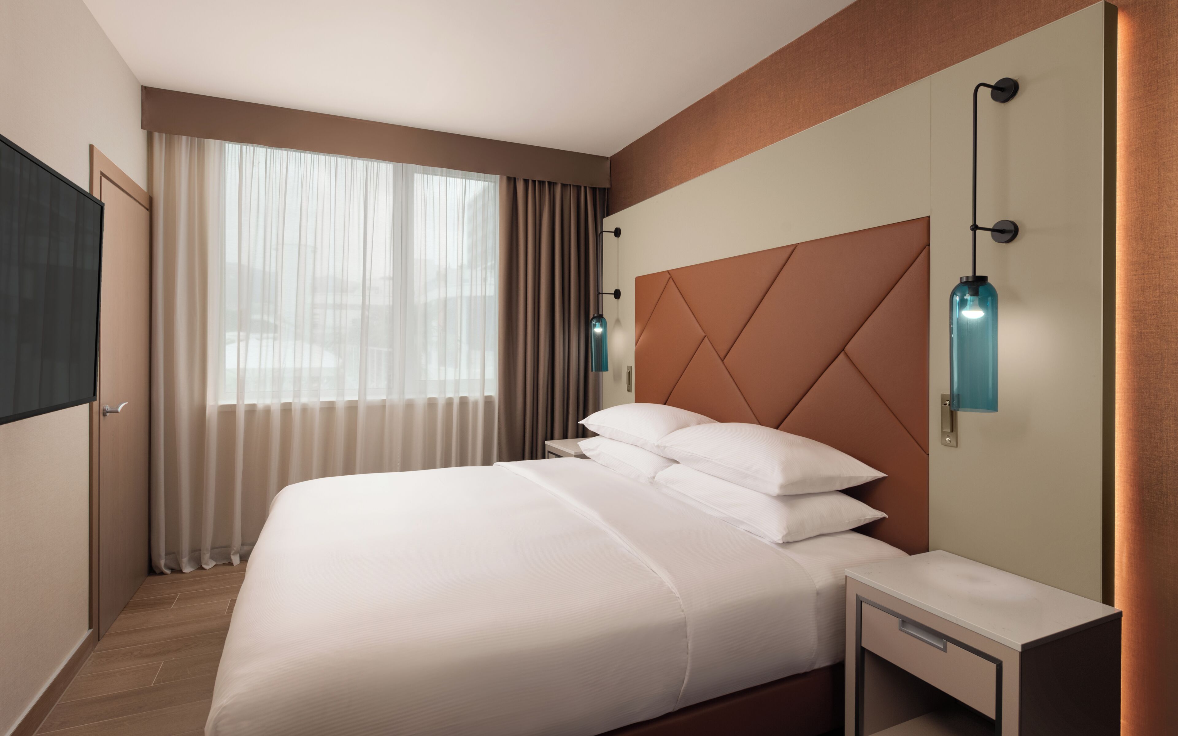 Quarto de hotel com cama, mesa de cabeceira, luminária na parede e janela com cortinas.