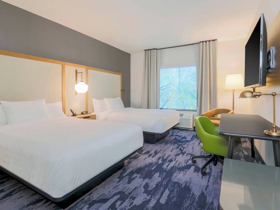Quarto de hotel com duas camas, uma escrivaninha, uma cadeira, uma televisão e uma janela com cortinas.