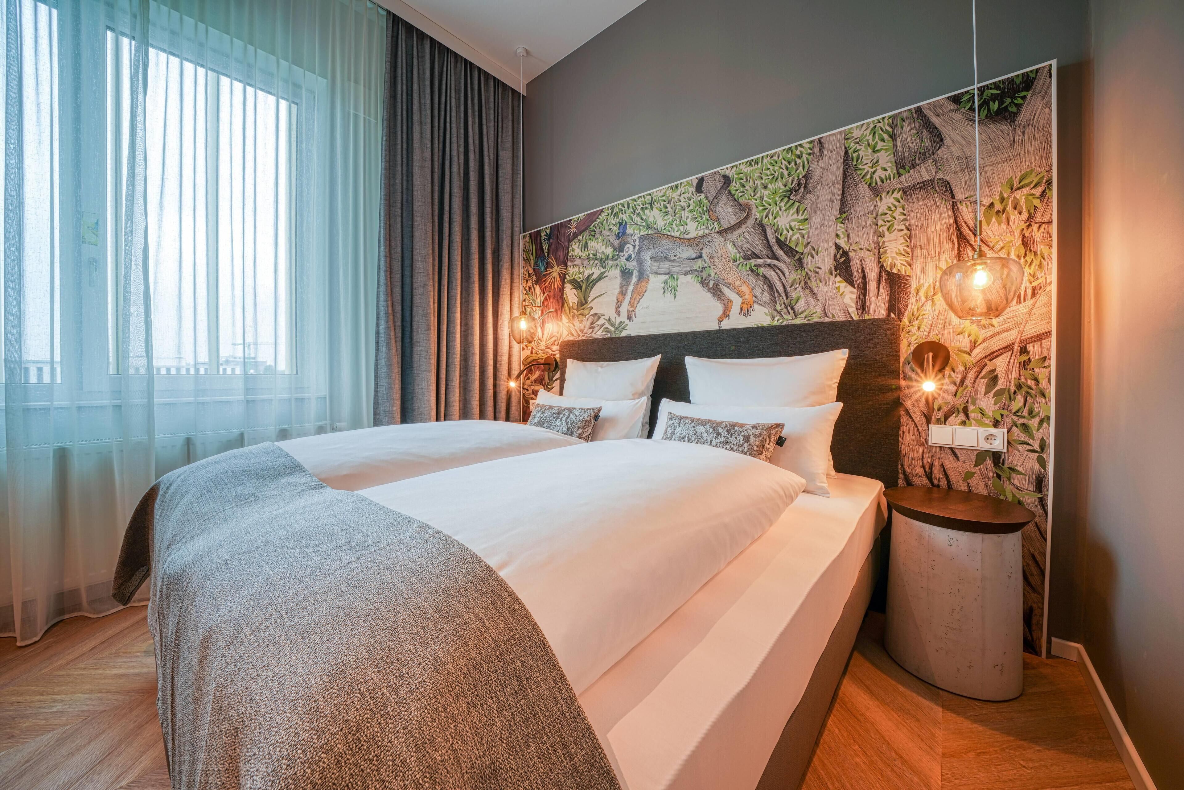Uma cama de casal com colcha em tons neutros, cabeceira com mural de tema natural, mesa de cabeceira redonda e janela com cortinas leves.