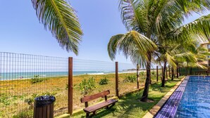 Área à beira da piscina com um banco, um cesto de lixo e uma cerca com vista para a praia.