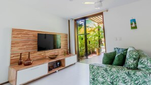 Uma sala de estar moderna com um rack de madeira, uma TV de tela plana, um sofá verde com padrão e uma porta de vidro deslizante que dá para um jardim tropical.
