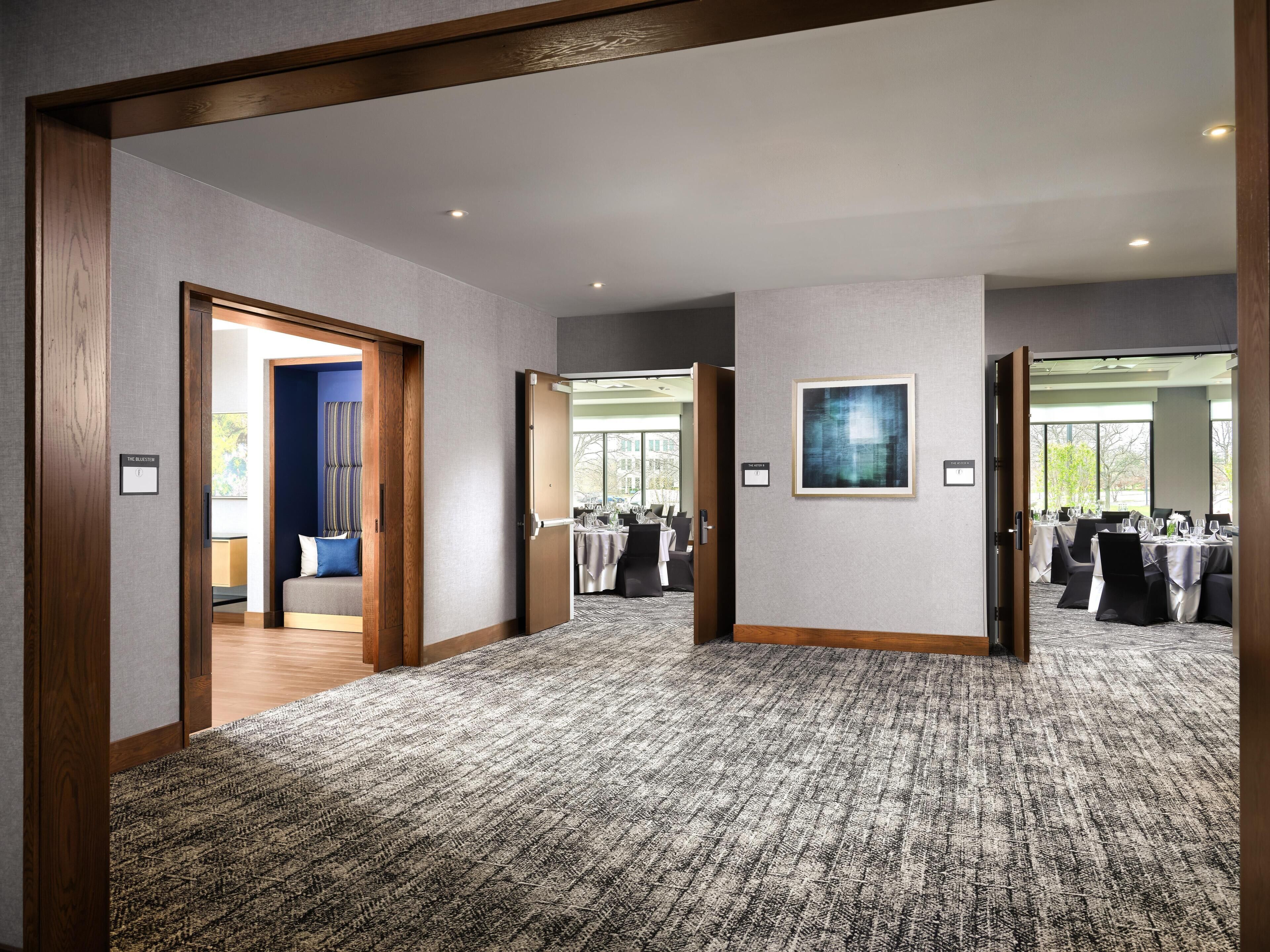 Um hall de hotel com piso revestido de carpete, molduras de porta em madeira e vista para uma sala de jantar com mesas postas.