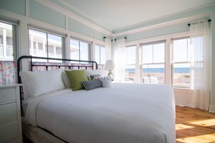 Quarto com cama grande, roupa de cama branca, mesa de cabeceira e vista para a praia através de grandes janelas.