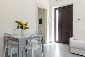 Uma área de refeições com uma mesa, cadeiras e um vaso de flores. Há uma porta e um interruptor de luz na parede.