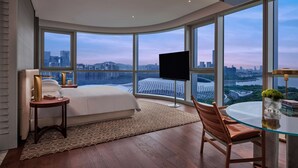 Quarto de hotel moderno com uma cama grande, televisão, mesa de vidro e vista para a cidade.
