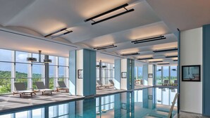 Piscina interior moderna com paredes de vidro transparente, espreguiçadeiras e vista para o exterior.