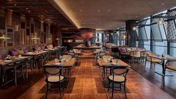 Um restaurante espaçoso com tetos altos, painéis de madeira e grandes janelas. Mesas montadas com cadeiras e centros de mesa.