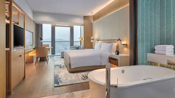 Quarto de hotel moderno com uma cama grande, área de estar com escrivaninha e cadeira, televisão, banheira e vista da cidade por amplas janelas.